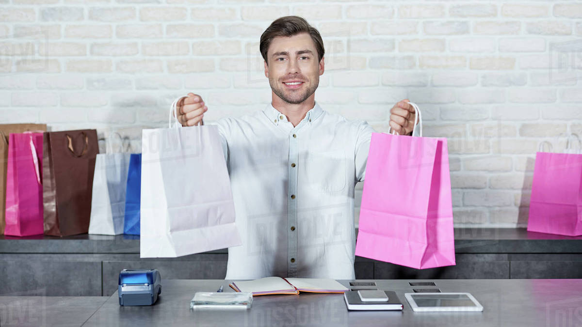 Handsome young salesman holding shopping bags and smiling at camera in ...