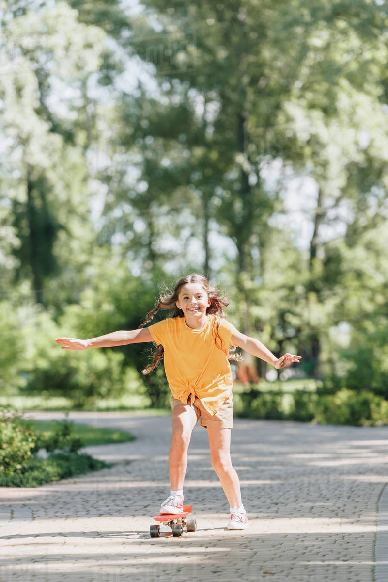Beautiful happy kid riding skateboard and smiling at camera in park ...