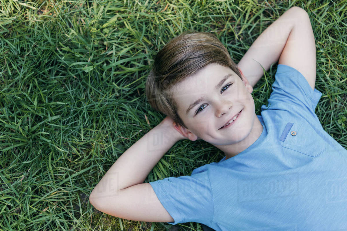 Top view of happy boy lying with hands behind head on grass and smiling ...