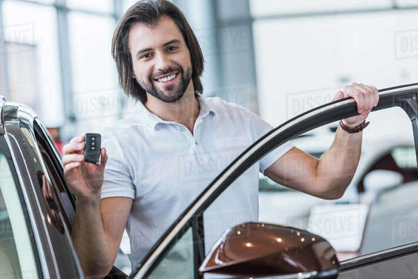 Portrait of happy man with car key standing at new car in dealership ...