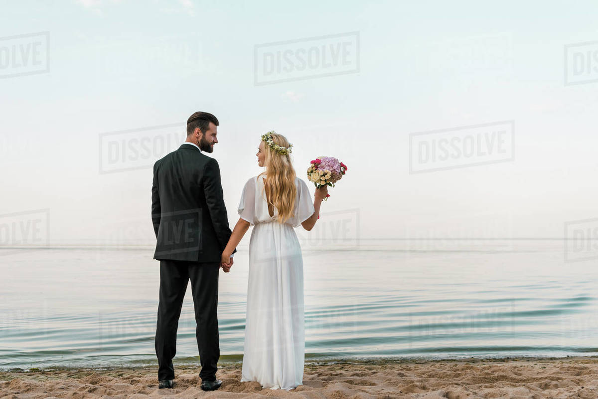 back view of wedding couple standing on beach with wedding bouquet and ...
