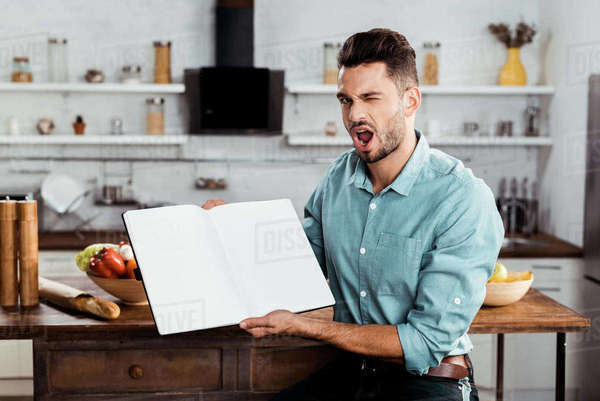 Handsome young man holding blank cookbook and winking at camera in ...