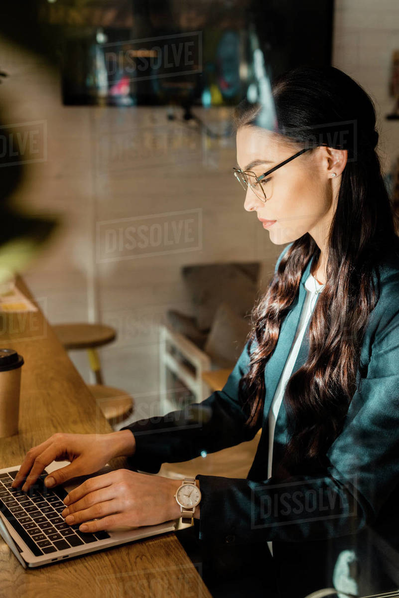 Beautiful brunette teleworker in eyeglasses using laptop at restaurant ...
