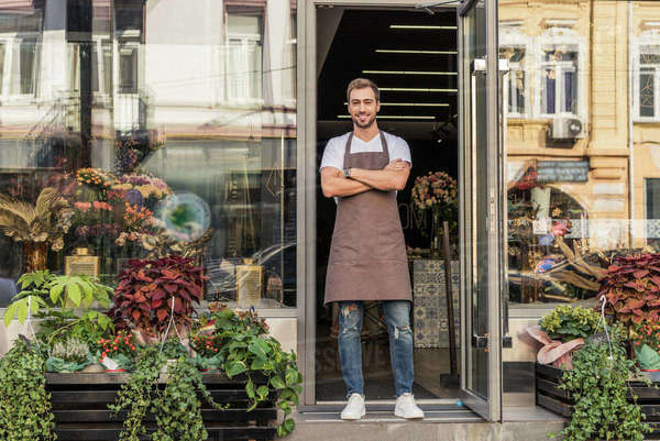 Smiling handsome florist standing near flower shop with crossed arms ...
