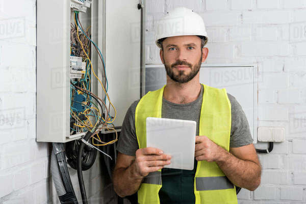 Handsome electrician holding tablet near electrical box in corridor and ...