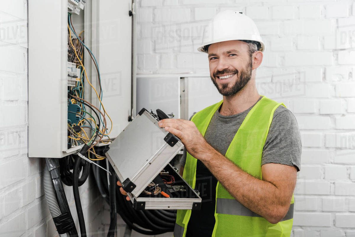 Smiling electrician holding toolbox near electrical box in corridor and ...