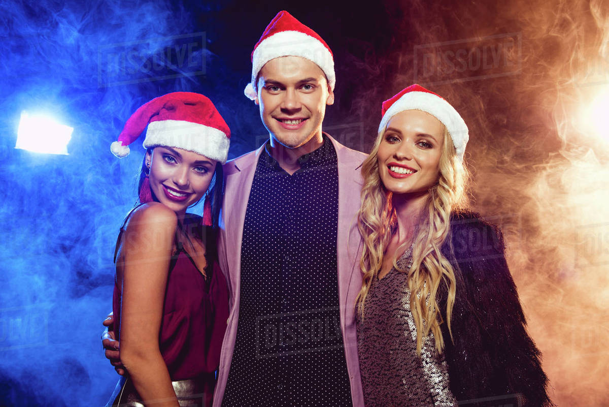 Smiling man in pink jacket embracing two women in Santa hats on smoke ...