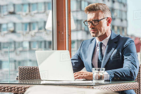 Focused businessman in eyeglasses using laptop at table with glass of ...