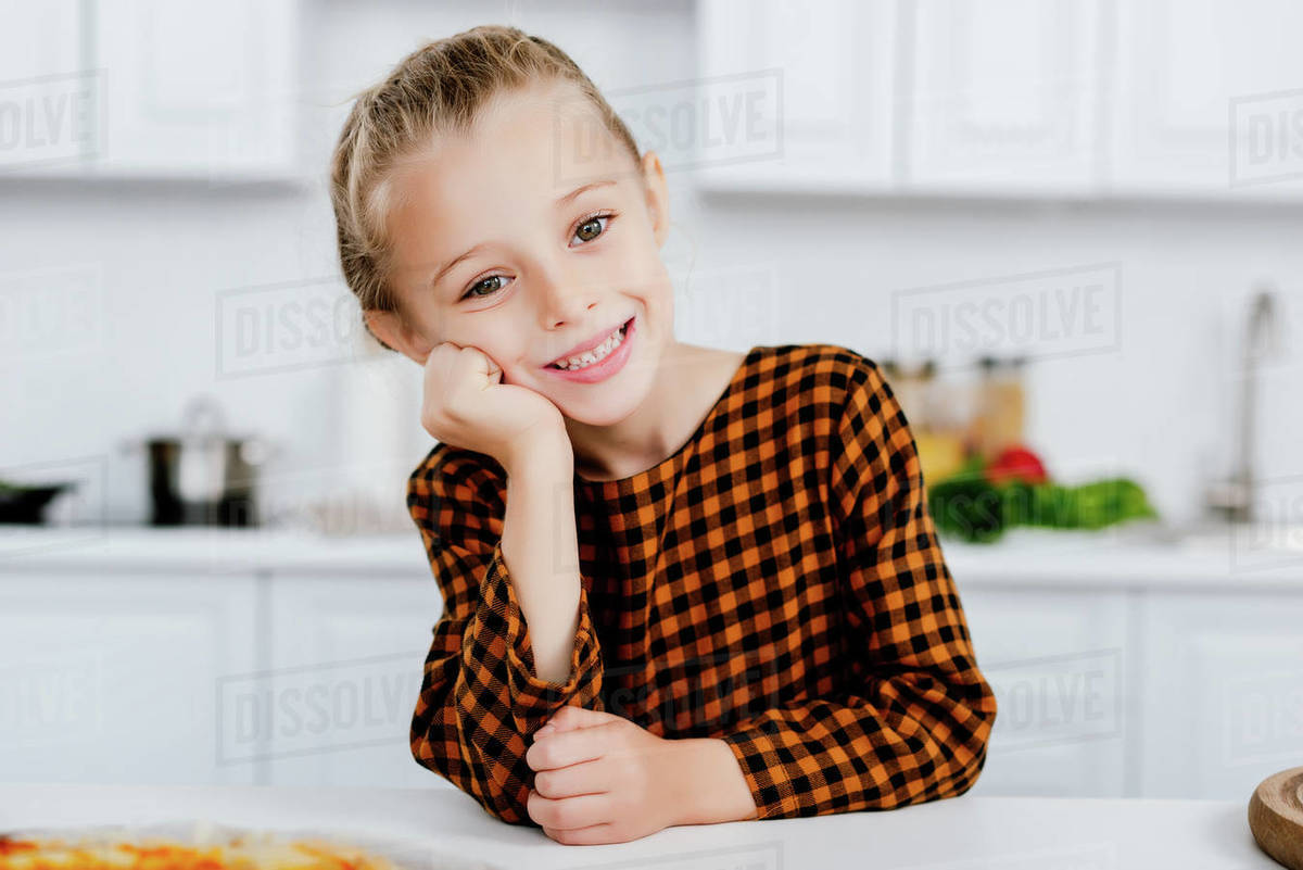 Adorable little child leaning on table at kitchen and looking at camera ...