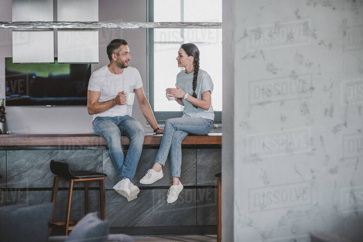 couple sitting on kitchen counter with cups of coffee in morning and ...
