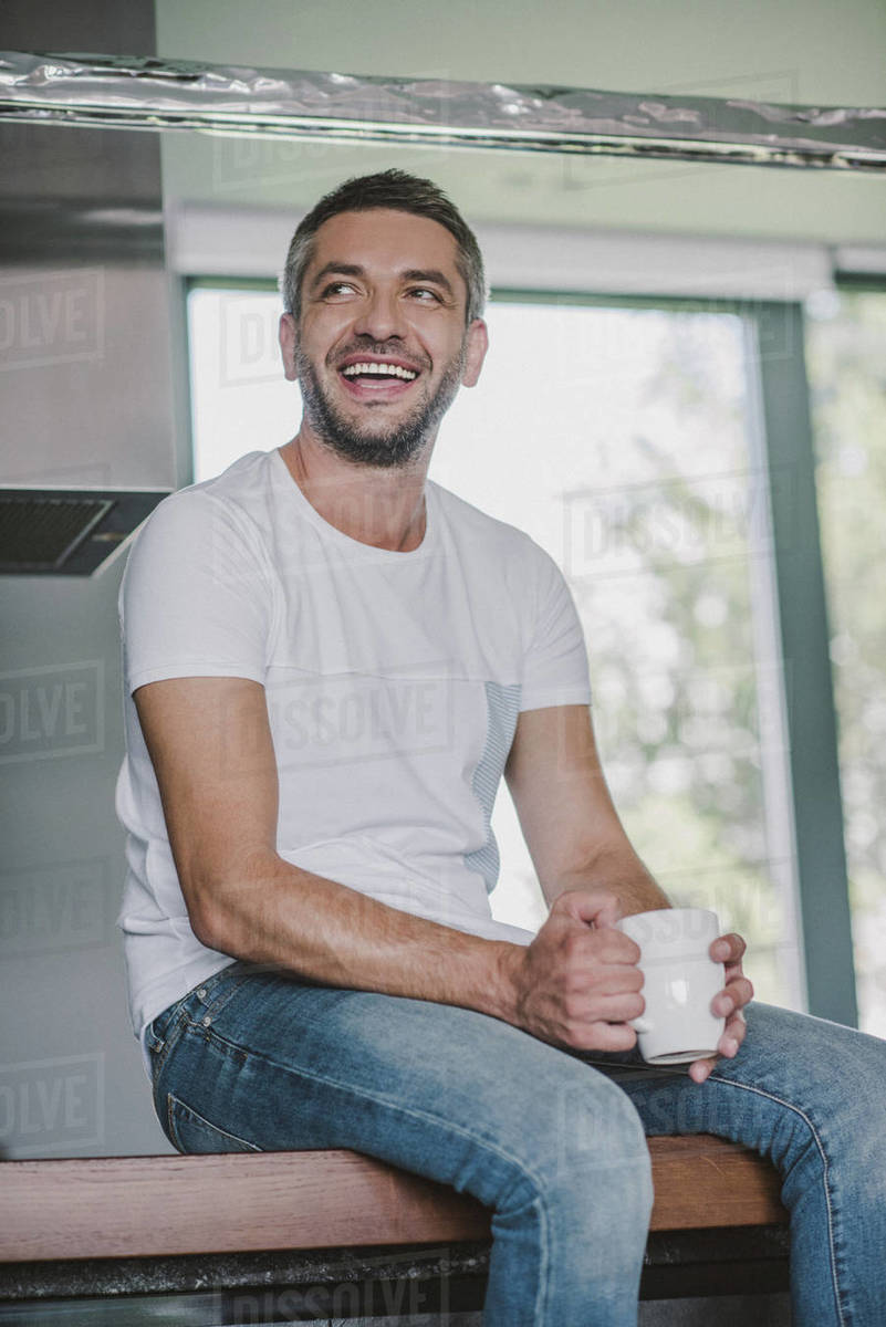 smiling handsome man sitting on kitchen counter with cup of tea in ...