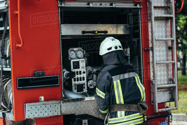 back view of firefighter in uniform and helmet standing at fire truck ...