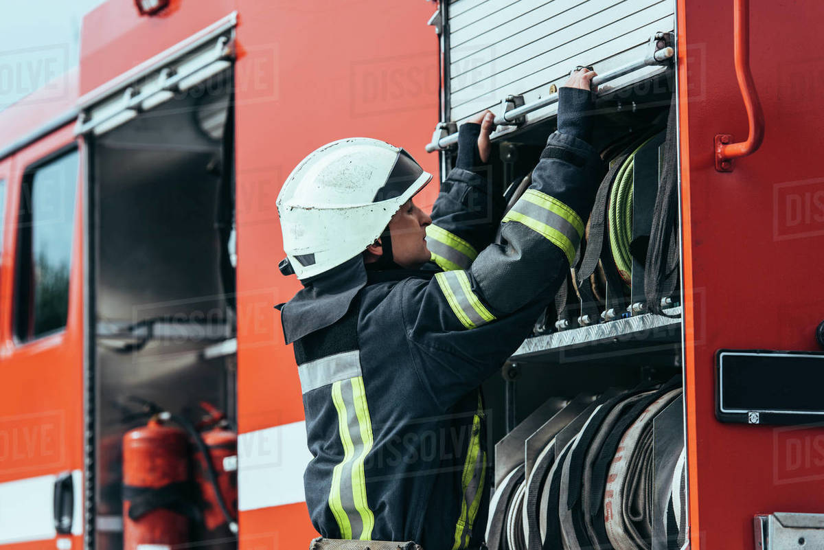 side view of fireman closing truck with arranged water hoses on street ...