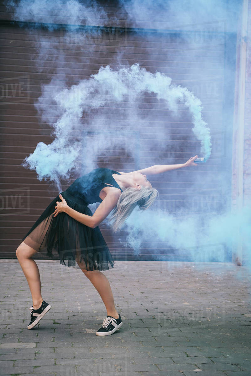 Side view of young female dancer dancing in blue smoke on street ...