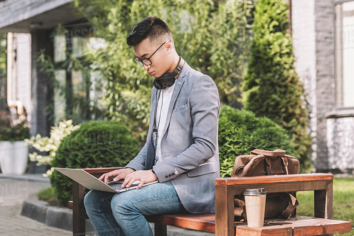 Asian teleworker using laptop while sitting on bench with leather bag ...