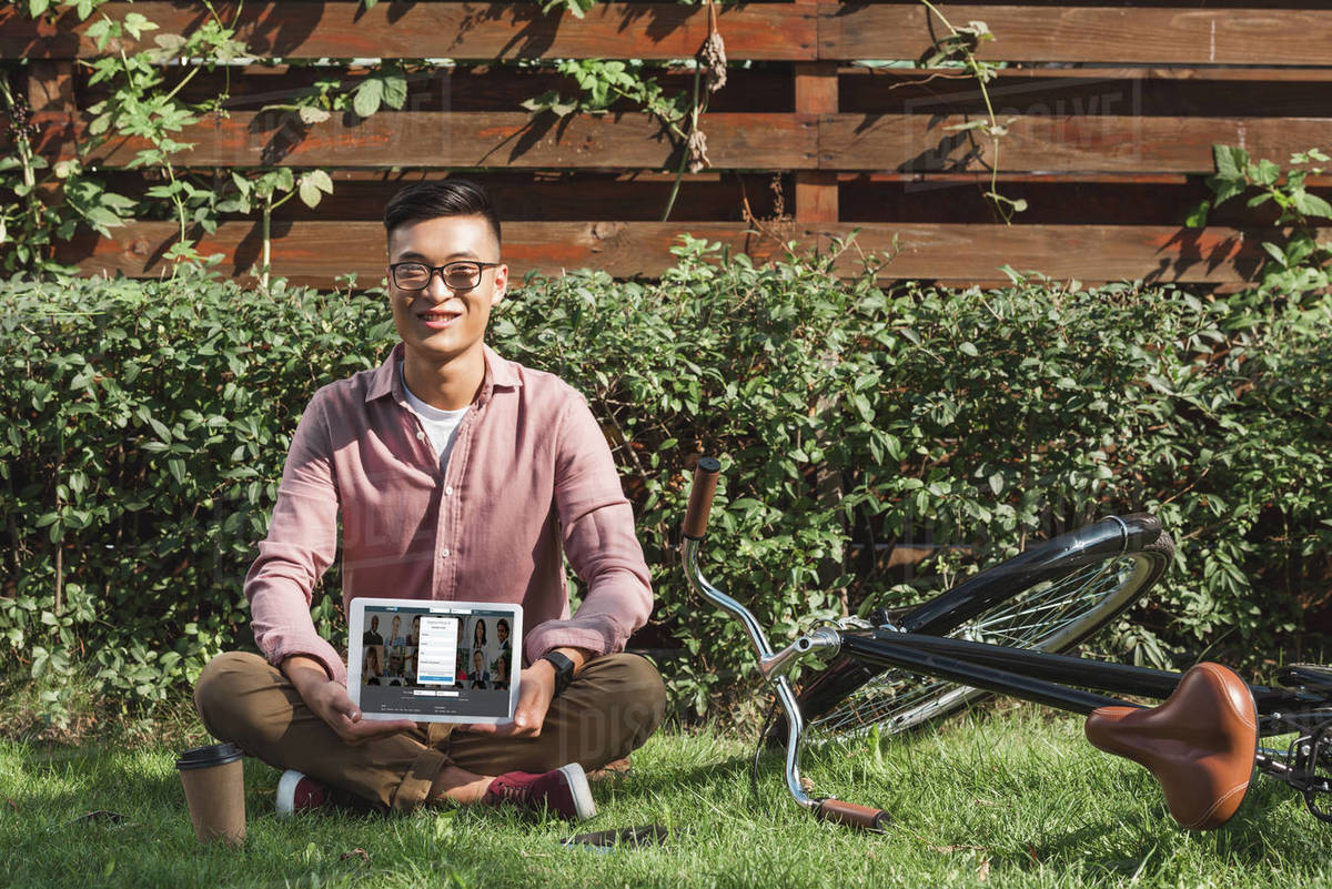 Smiling Asian man sitting on green grass and showing tablet with ...