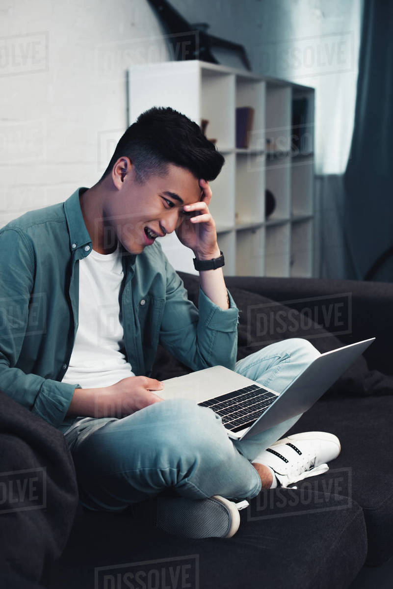 Smiling young Asian man sitting on couch and using laptop at home ...