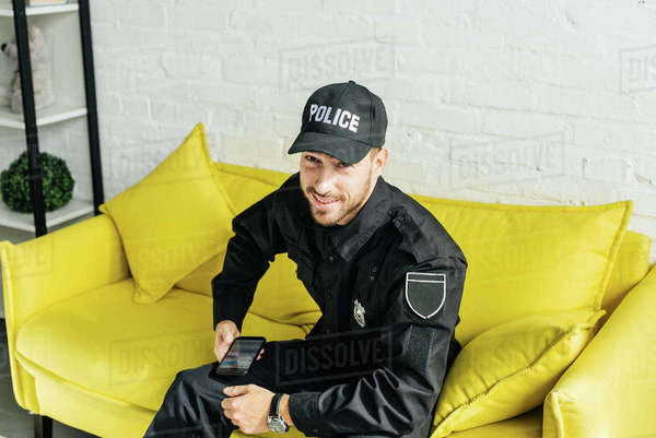 Happy young policeman with smartphone sitting on yellow couch and ...