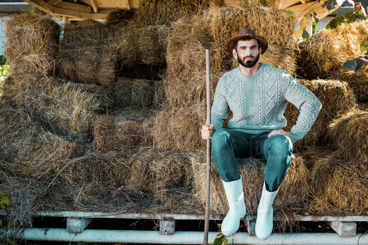 Handsome male farmer in sweater and straw hat sitting on hay stacks at ...