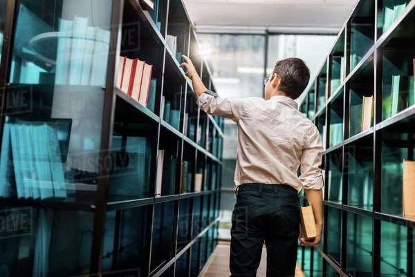 Back view of boy holding book and looking at bookshelves in library ...