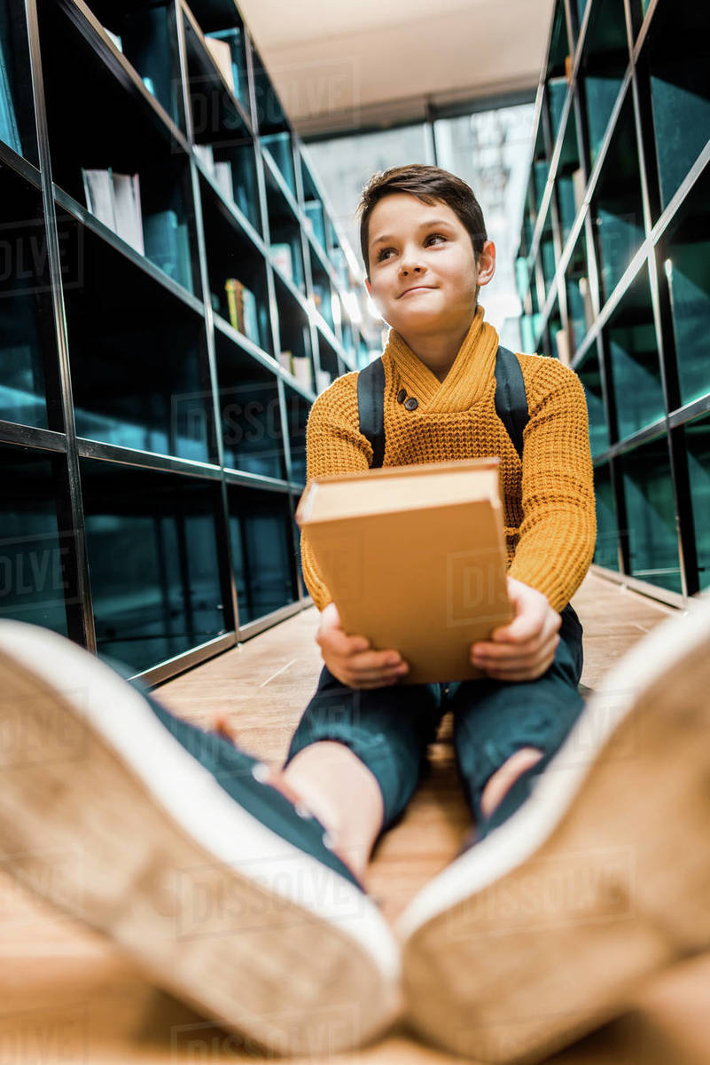 Adorable smiling schoolboy holding book and sitting on floor in library ...