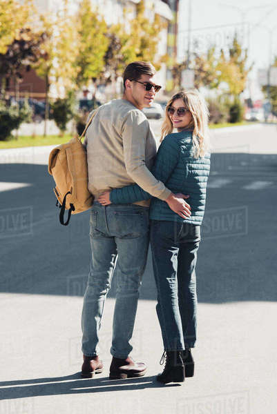 Back view of couple hugging and looking at camera while walking on ...