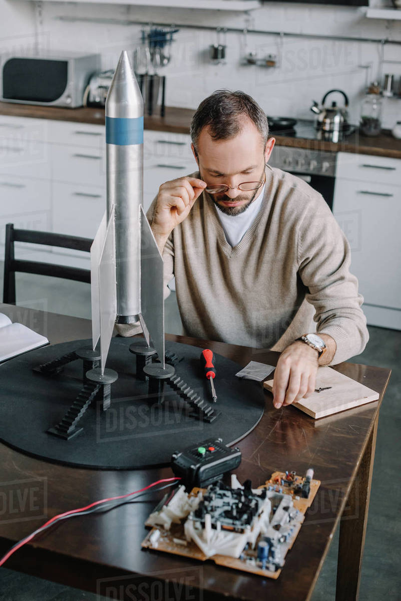 Handsome engineer modeling rocket and looking above glasses at table at ...