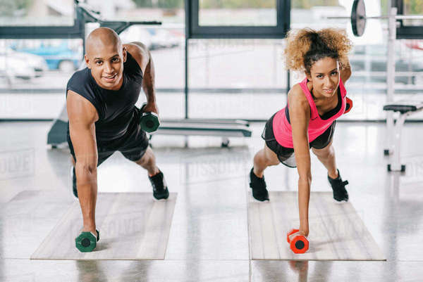 Cheerful African American couple of athletes exercising with dumbbells ...
