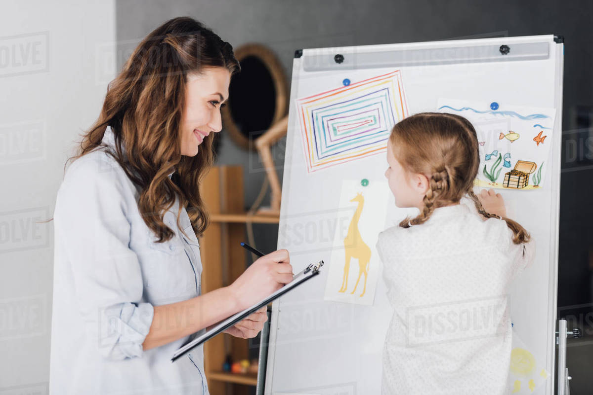Happy psychologist with clipboard talking to child near whiteboard with ...