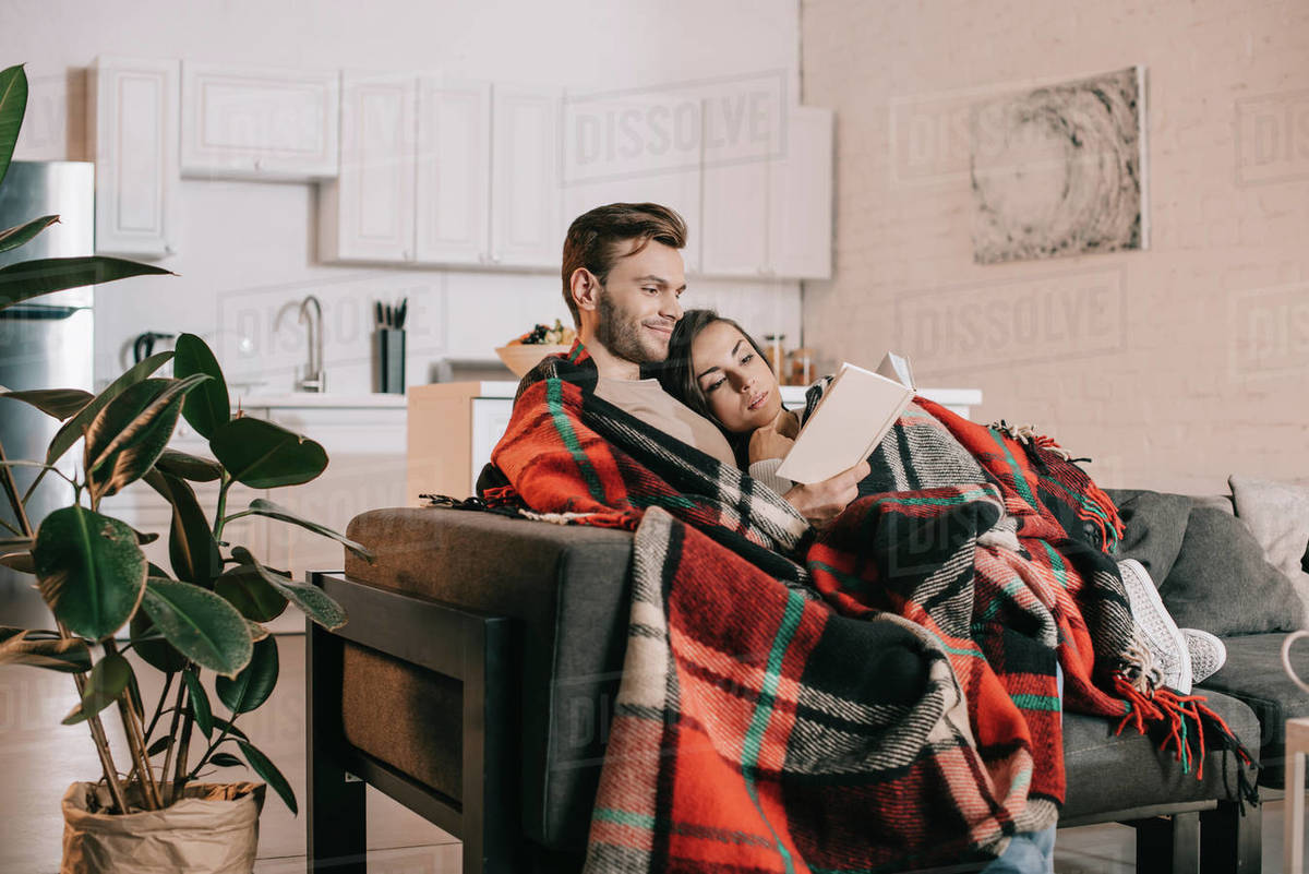 Smiling young couple reading book together on couch under plaid at home ...
