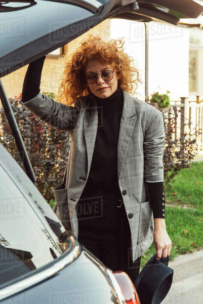 Stylish curly redhead woman in grey jacket and eyeglasses closing car ...