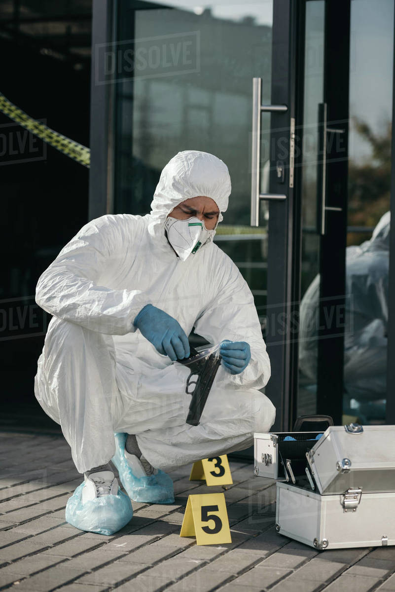 Male criminologist in protective suit and latex gloves sitting with evidence gun in arms ...
