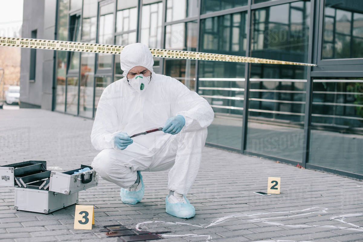 Male criminologist in protective suit and latex gloves explore the murder weapon at crime scene ...
