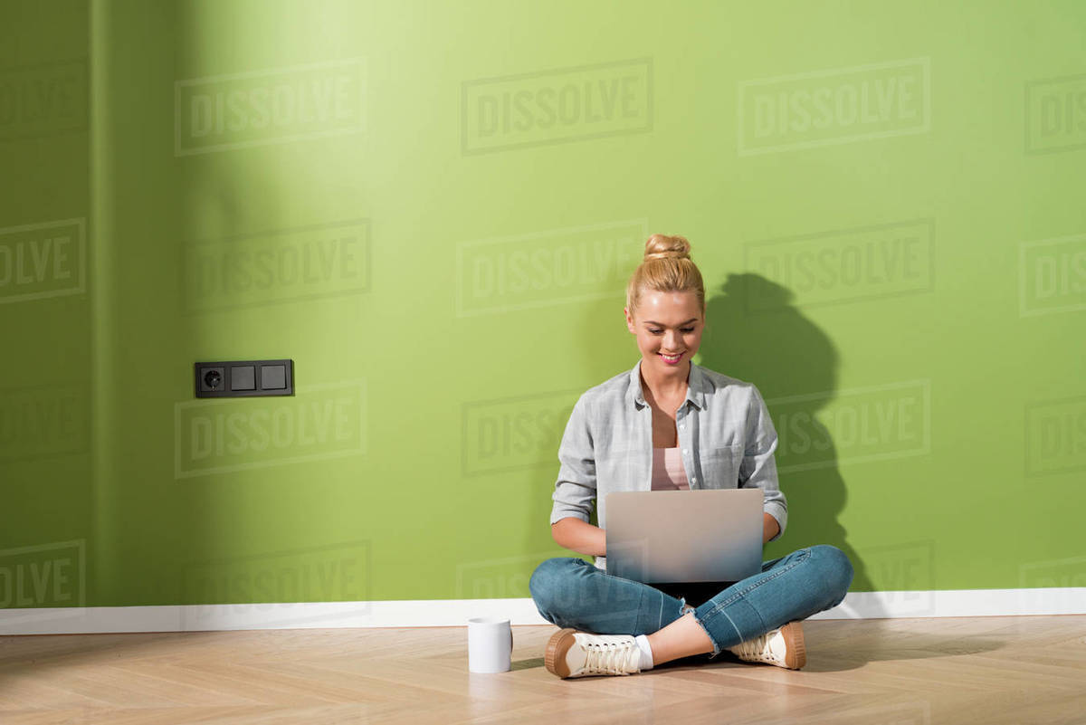 Smiling girl typing on laptop keyboard and sitting on floor by green ...