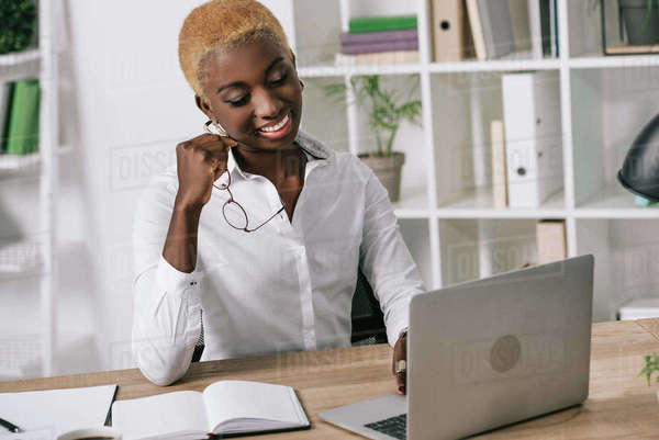 African American businesswoman smiling and holding glasses near laptop ...
