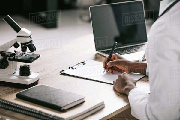 Cropped view of African American scientist writing on paper near laptop ...