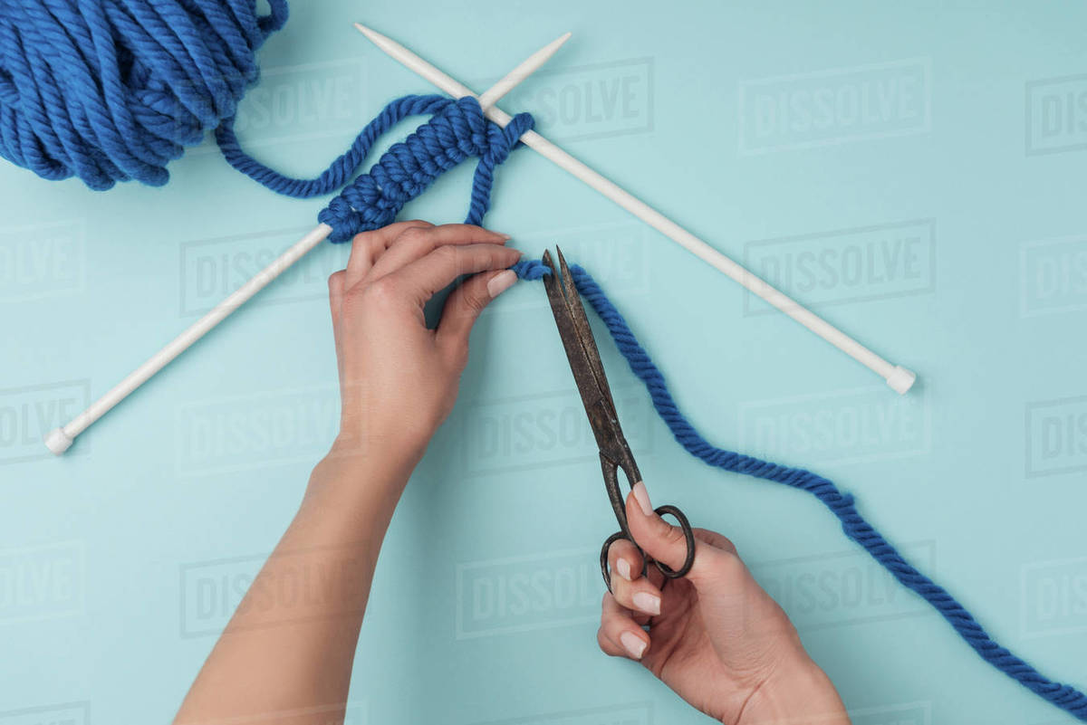 partial view of woman cutting thread with scissros on blue backdrop ...