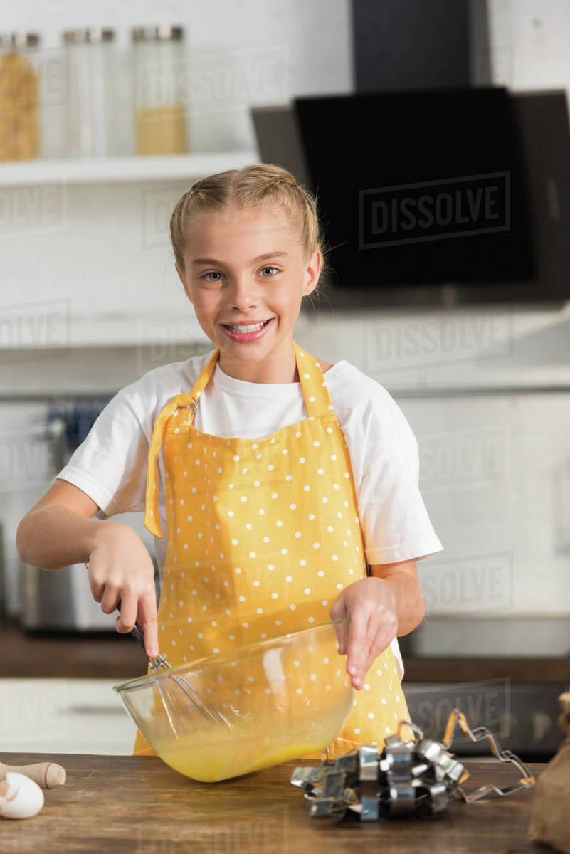 Adorable happy child in apron whisking eggs and smiling at camera in ...