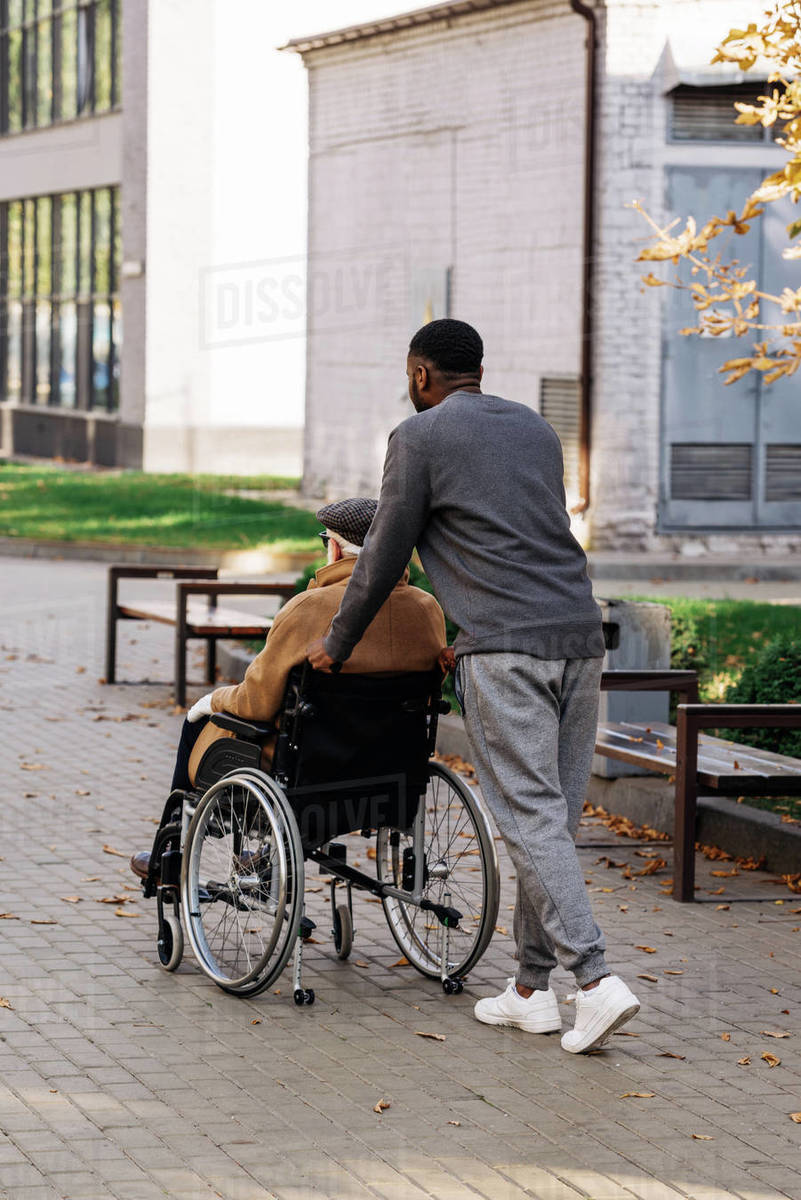 Rear view of senior disabled man in wheelchair and nurse riding on ...
