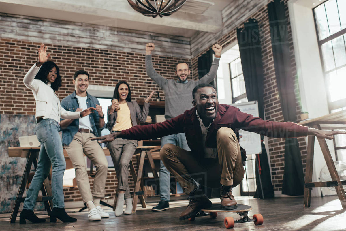Group of happy multiethnic coworkers having fun with skateboard in loft ...
