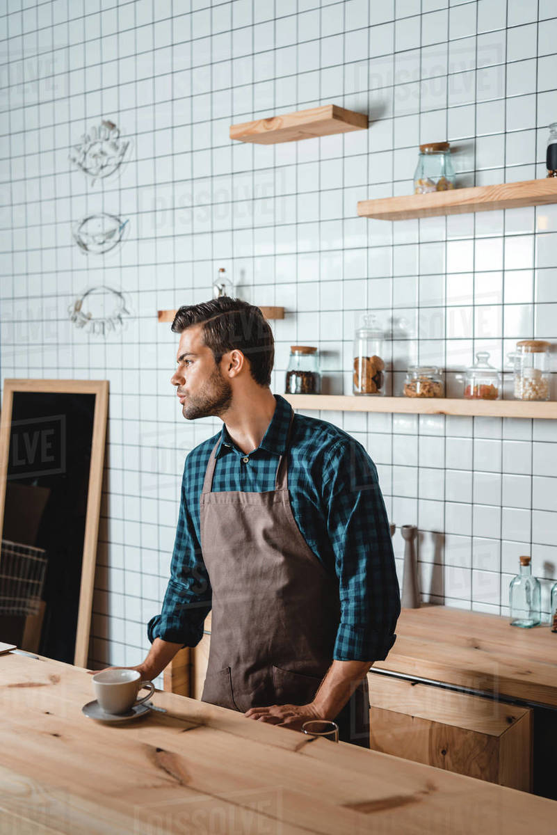 side view of pensive barista standing at counter with cup of coffee in ...