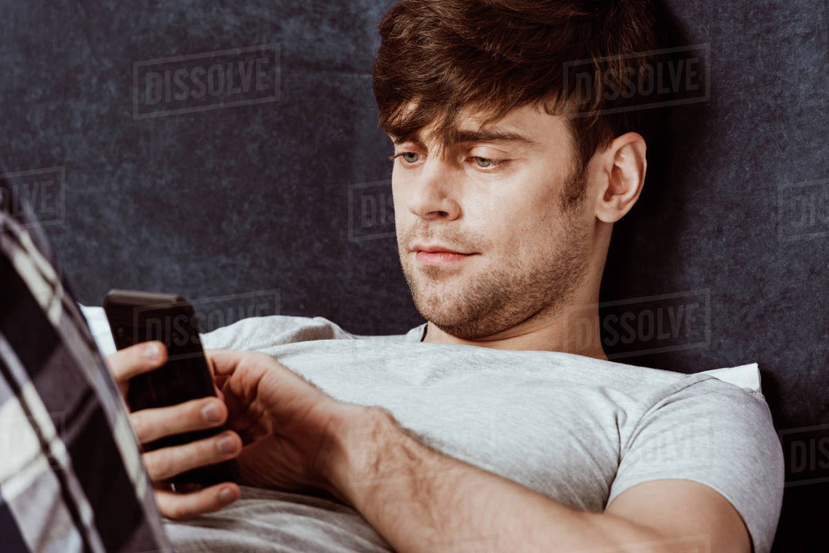 portrait of handsome man using smartphone while laying in bed at home ...