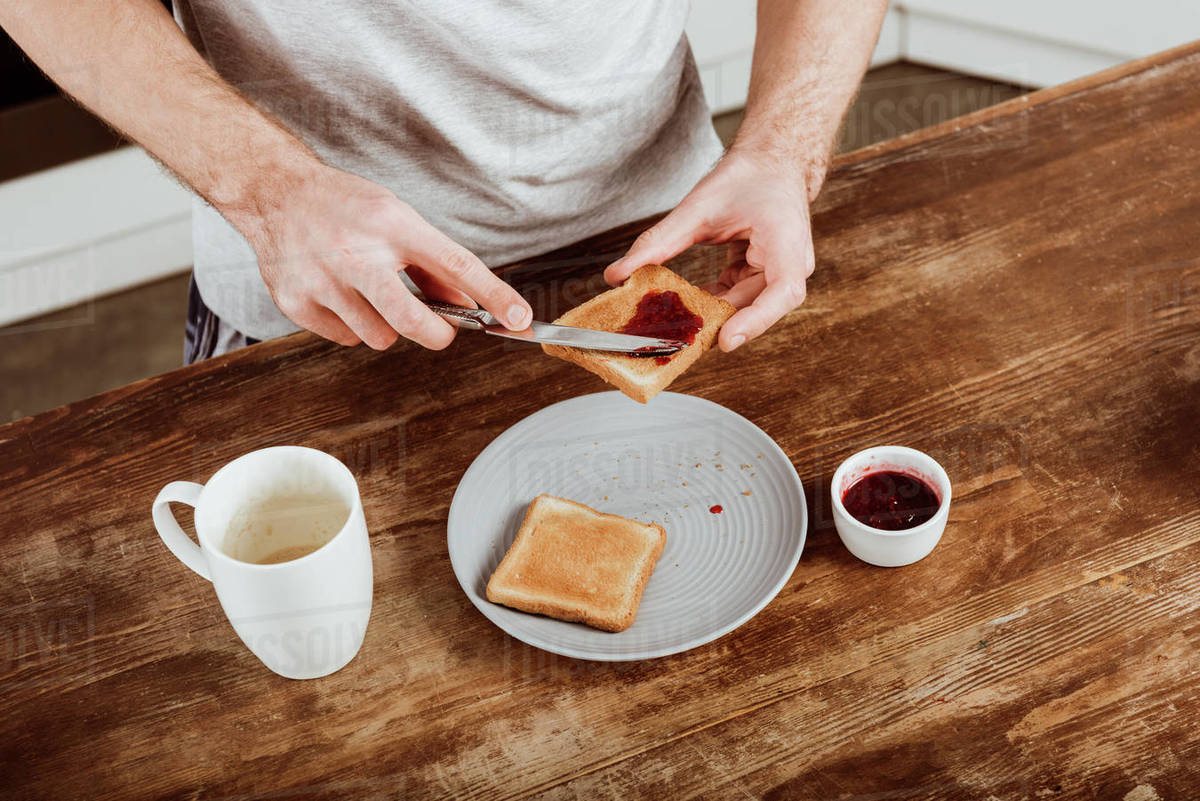 partial view of man spreading toast by jam at table with coffee cup in ...