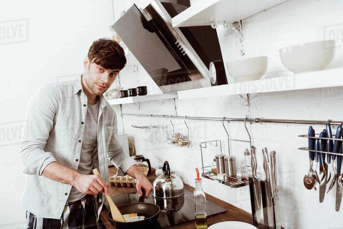 selective focus of young man cooking scrambled eggs on stove in kitchen ...