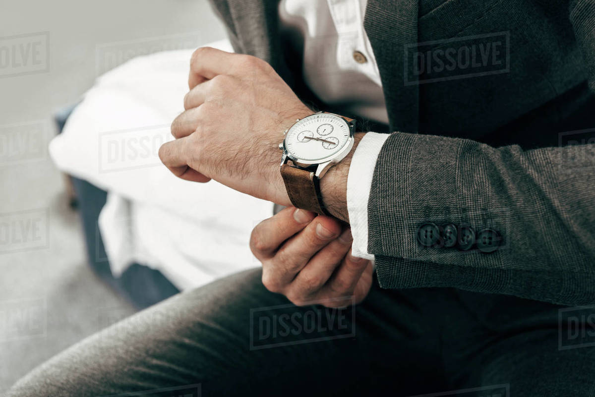 partial view of businessman in jacket putting on hand watch at home ...