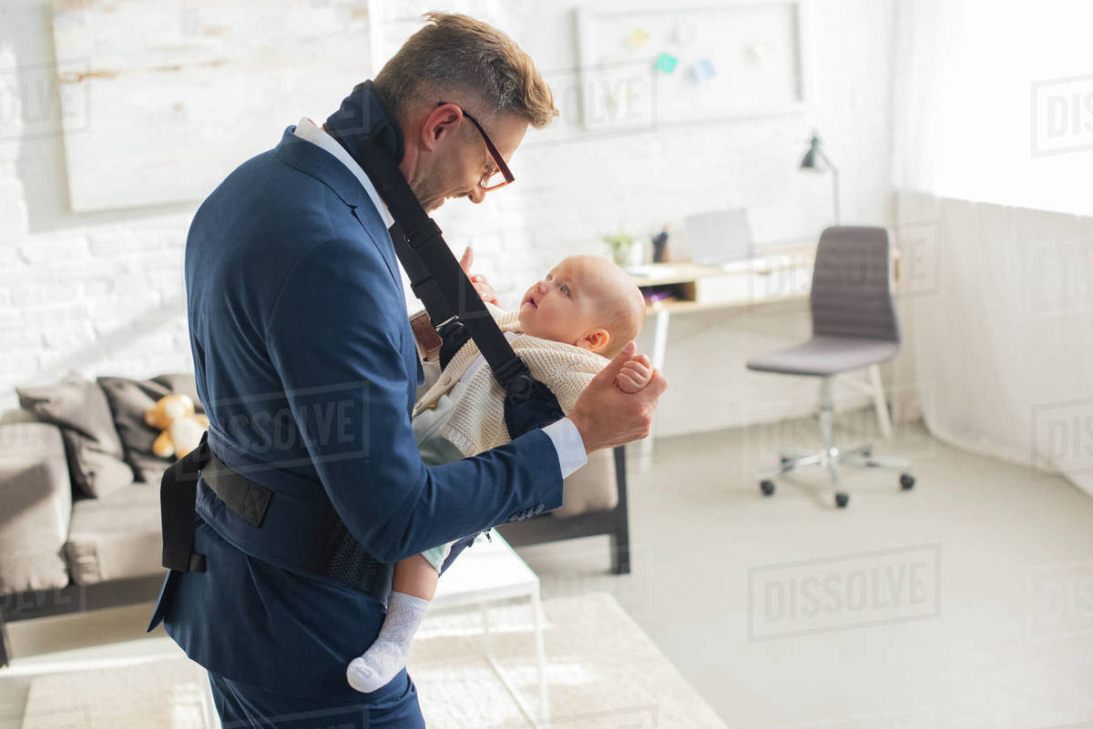 Cheerful businessman holding hands of infant daughter in baby carrier ...