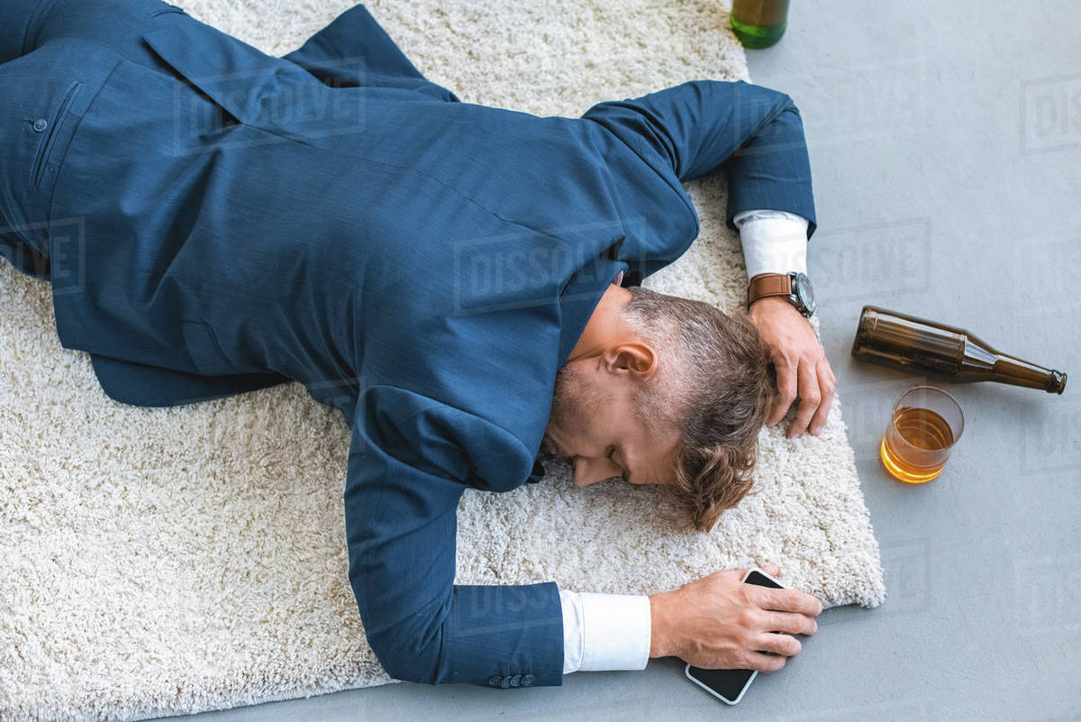 Top view of drunk businessman lying on carpet with smartphone in hand ...