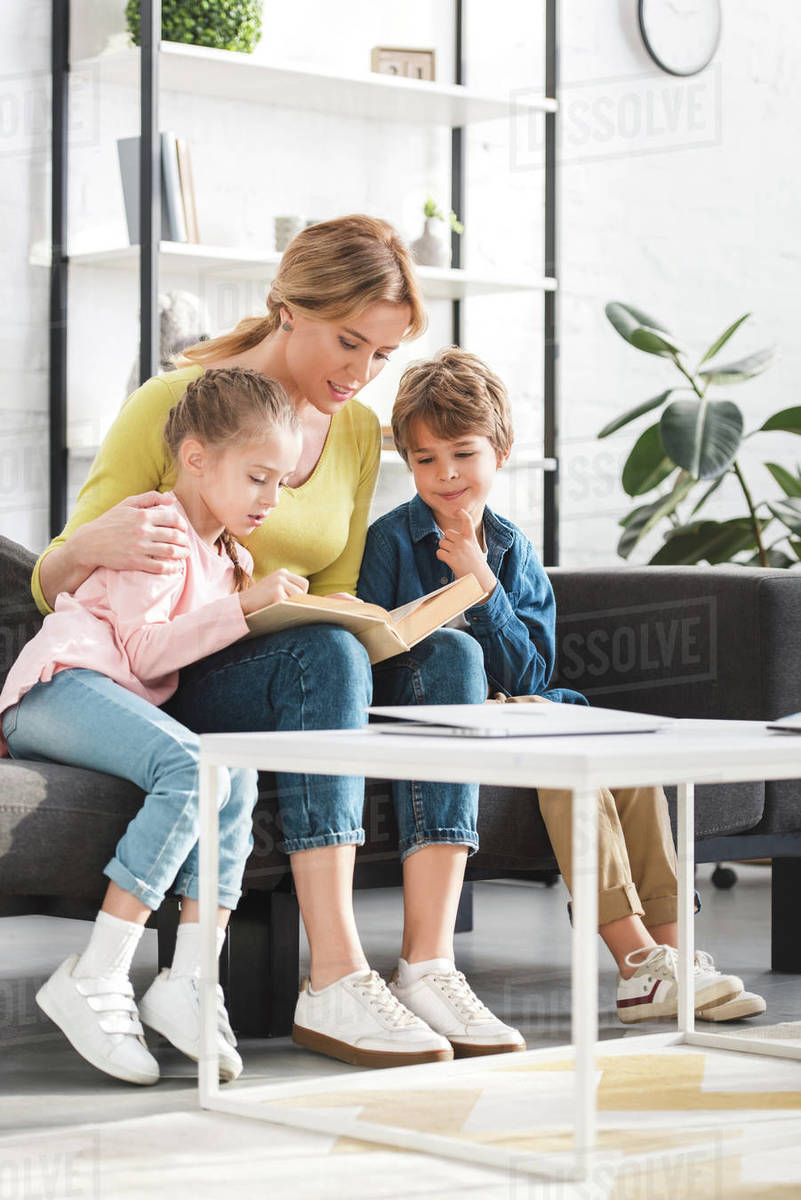 Mother with adorable children sitting on sofa and reading book ...