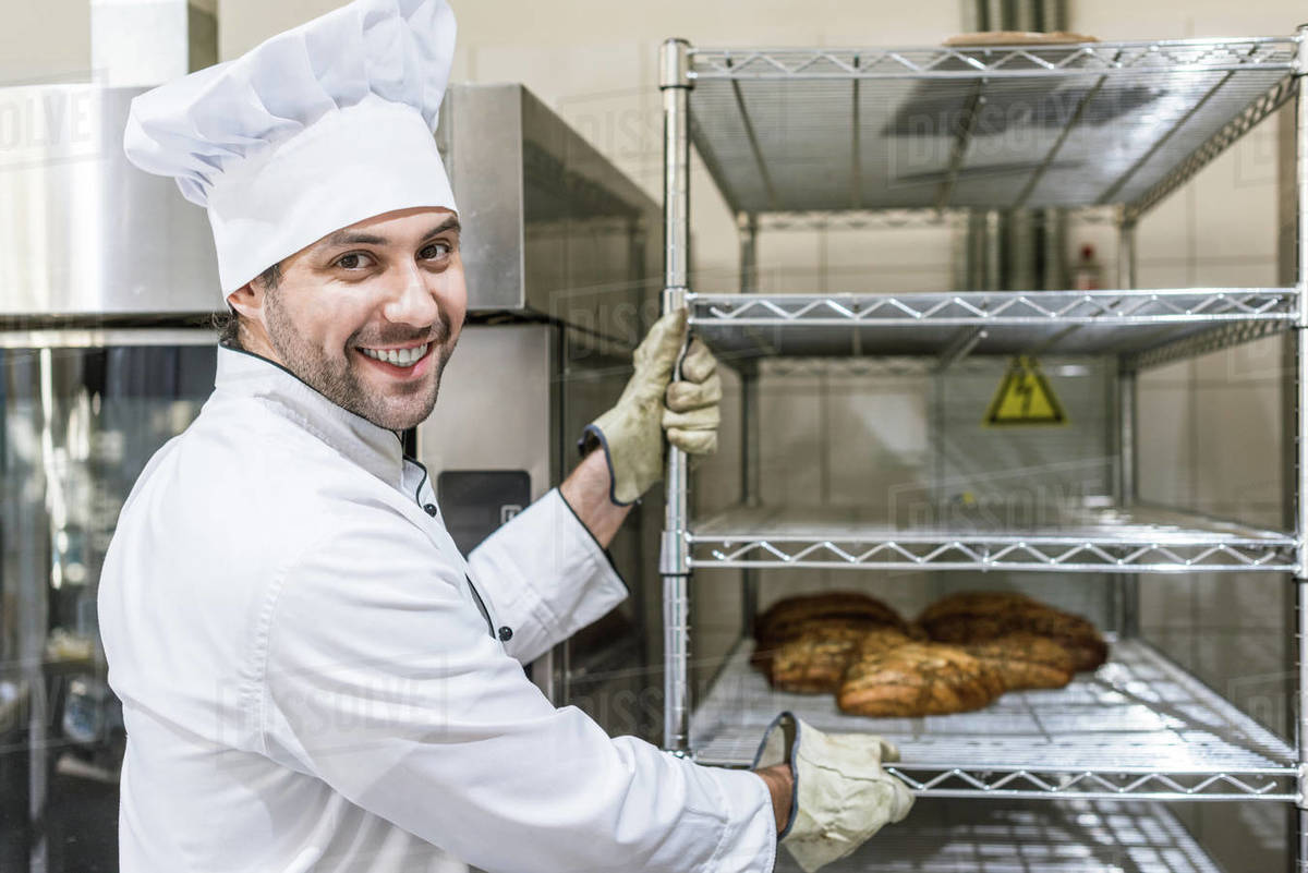 smiling male baker with hot baked bread on rack - Stock Photo - Dissolve