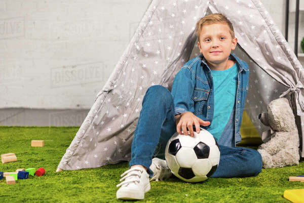 positive little boy sitting with soccer ball near tent at home ...